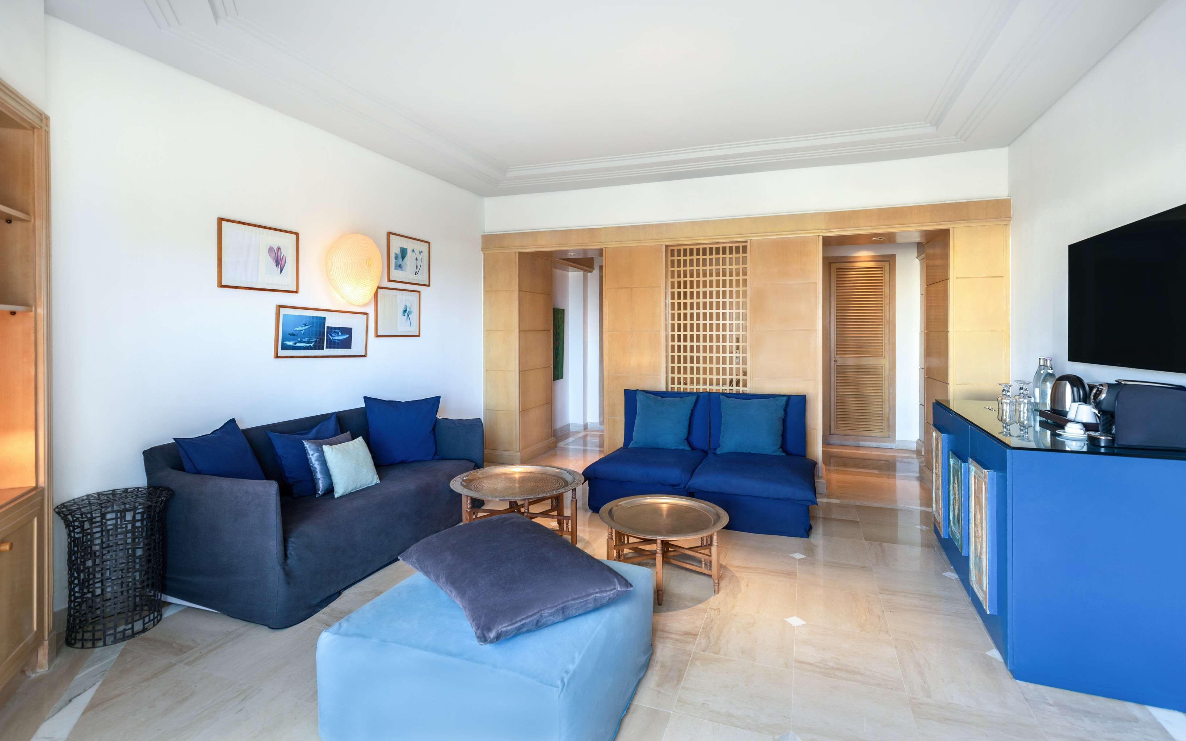 Uma sala de estar moderna com um sofá azul escuro, um banquinho azul claro e uma mesa de centro. Há uma área de cozinha com bancada azul e uma TV de tela plana.