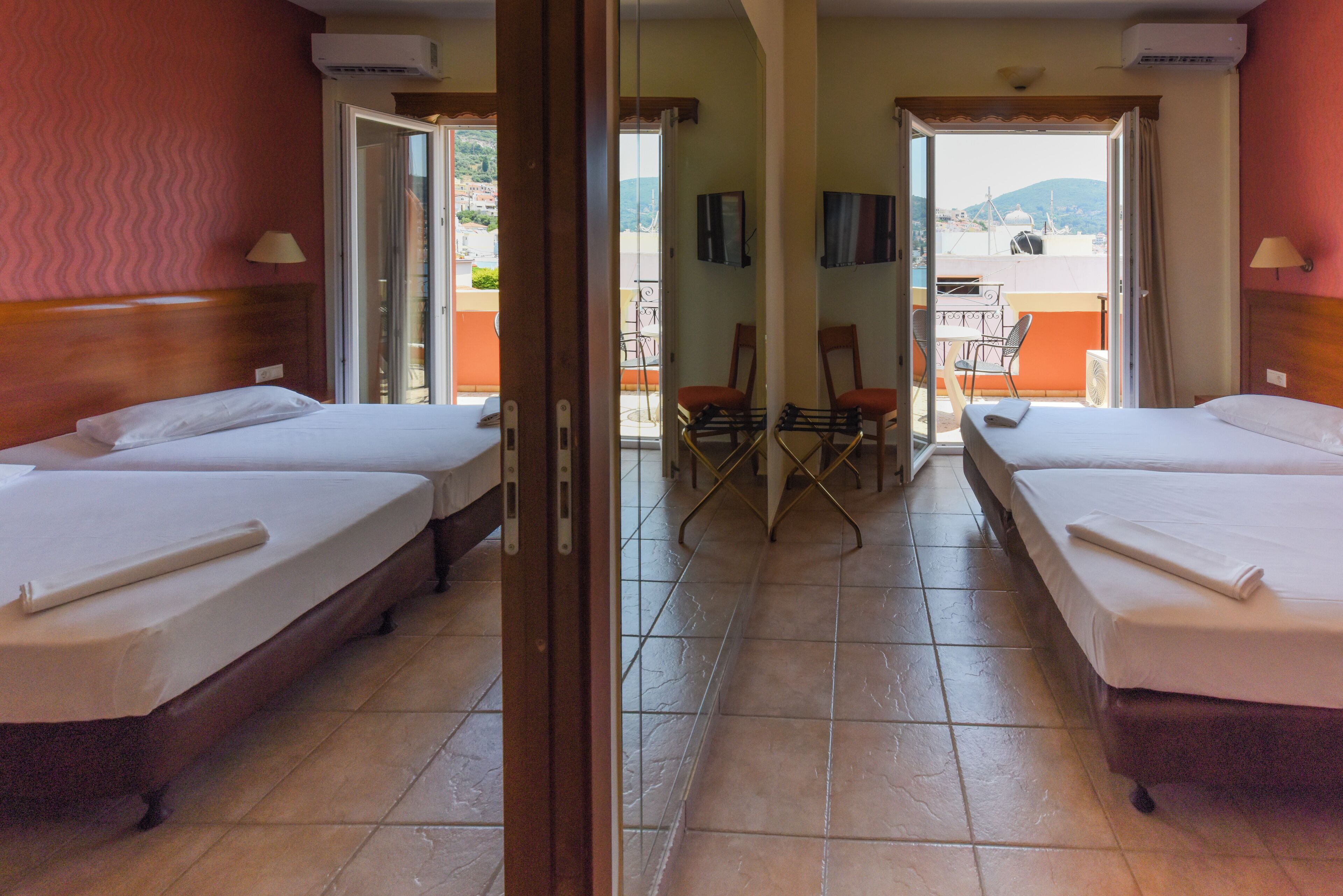 Quarto de hotel com duas camas, varanda com mesa e cadeiras, e vista para a paisagem urbana.