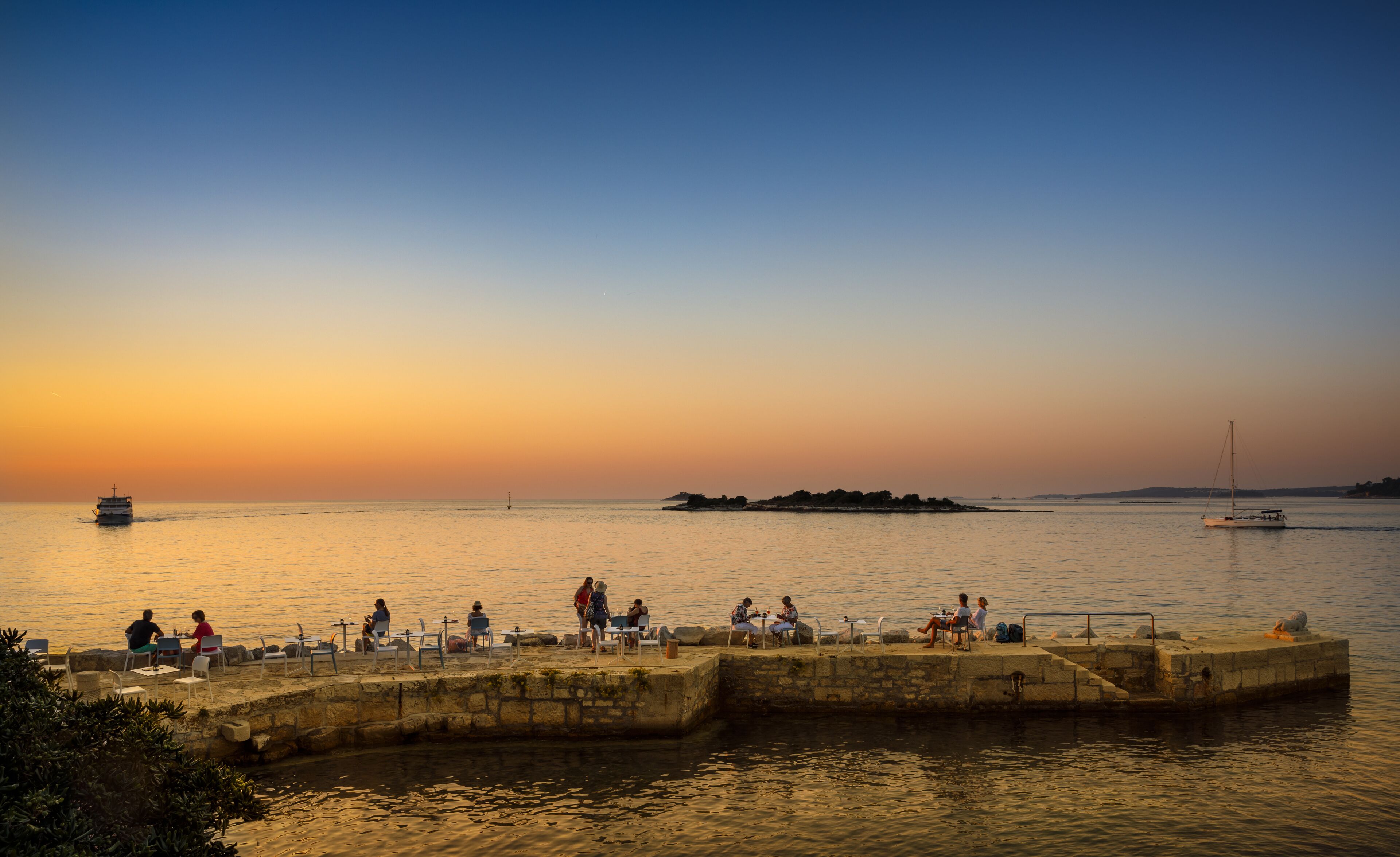 Um cenário à beira-mar com um cais, pessoas sentadas em mesas e barcos na água.
