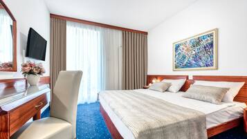 Quarto de hotel com cama, escrivaninha, cadeira, televisão e um quadro na parede.