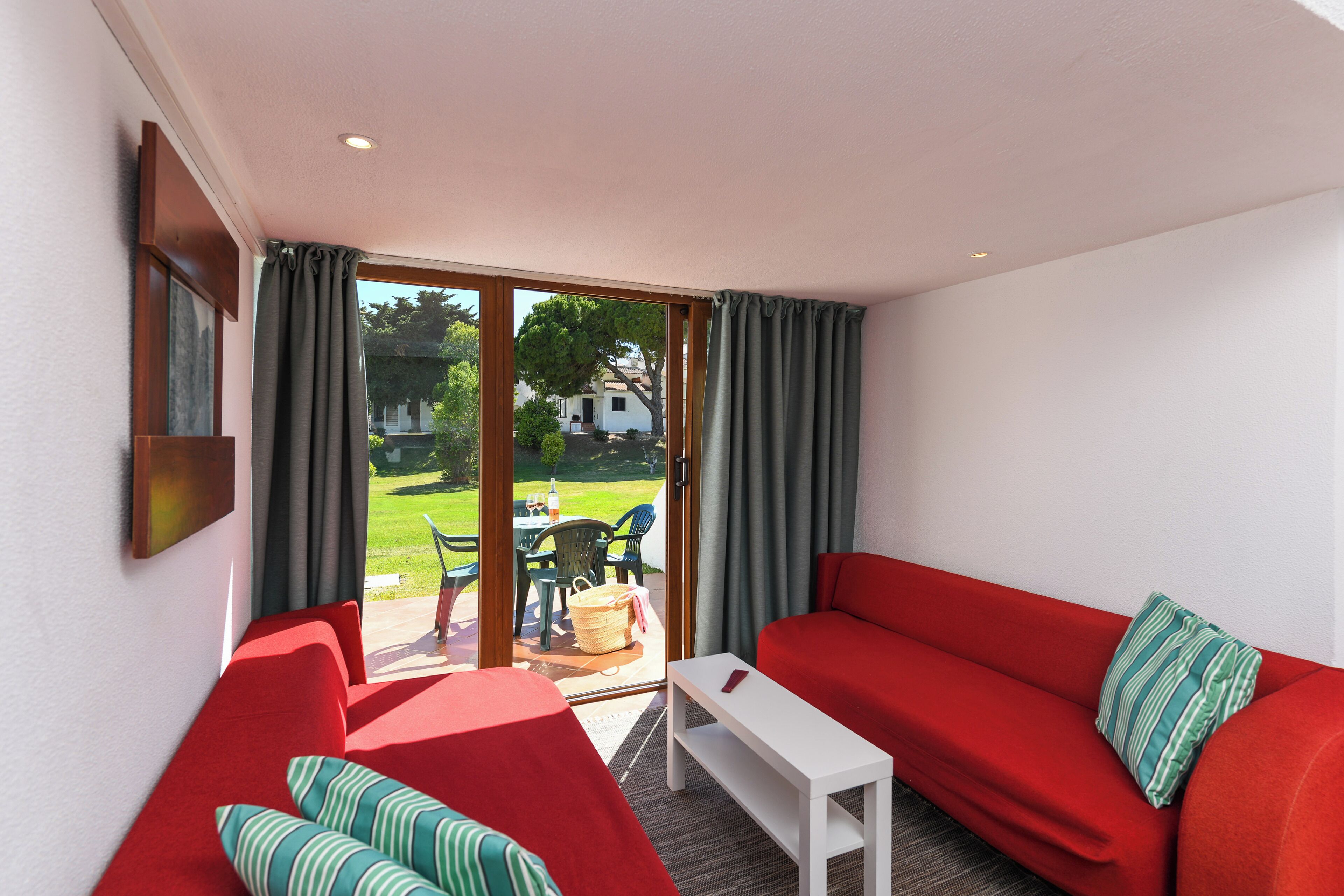 Um sofá vermelho, uma mesa branca e vista para um jardim através de uma porta de vidro.