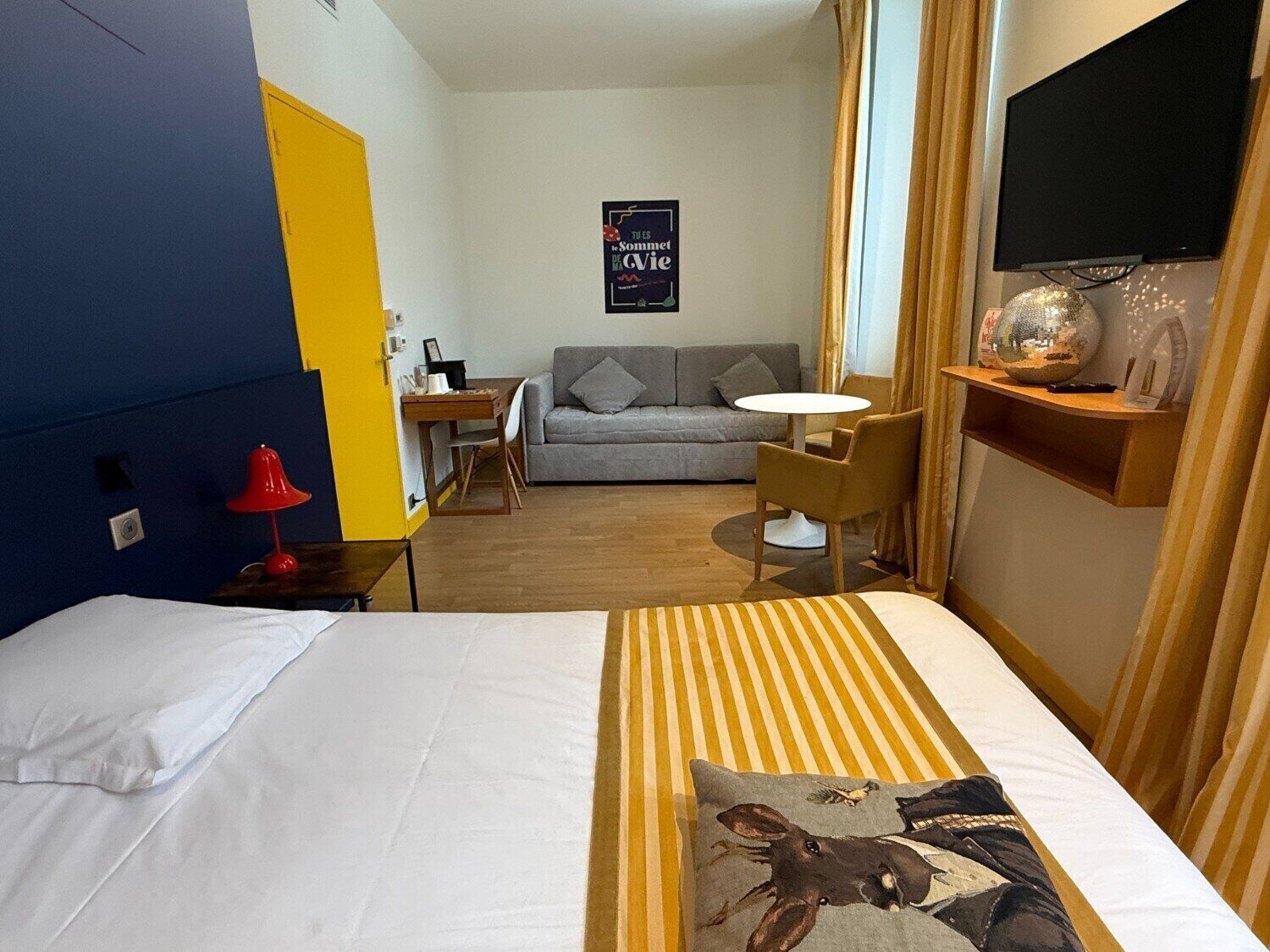 Quarto de hotel com cama, sofá, mesa pequena, televisão e um cartaz na parede.