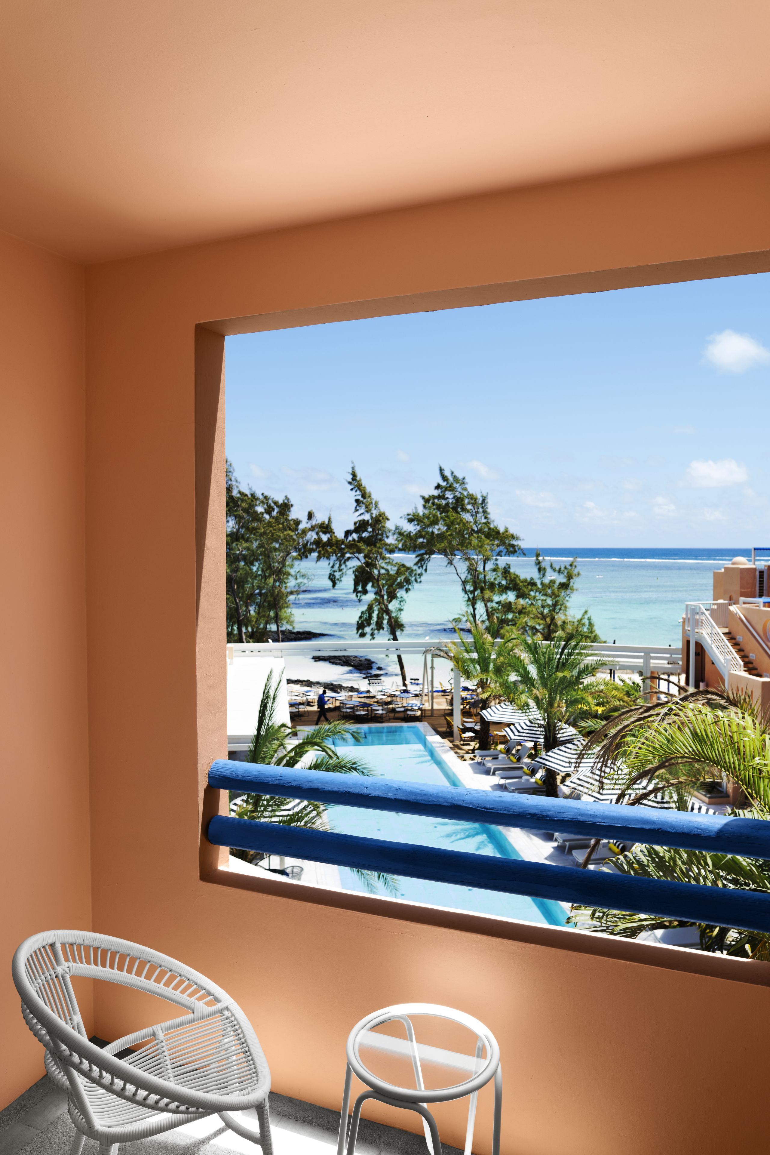 Varanda com vista para uma piscina e praia, cadeiras de vime branco e uma pequena mesa.