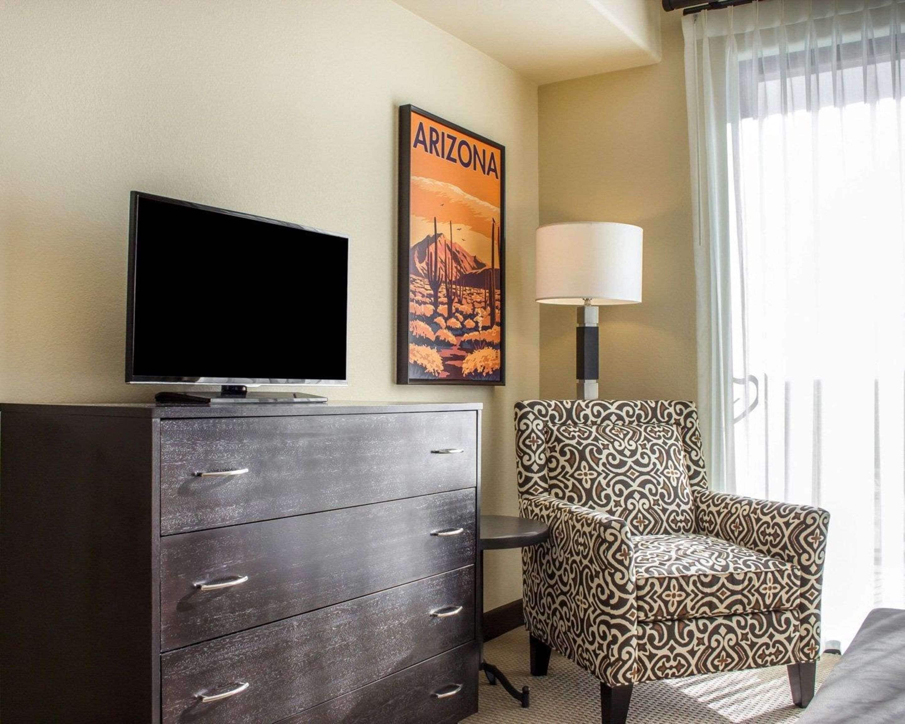 Quarto de hotel com cómoda de madeira escura, televisão, poltrona estampada, luminária e um cartaz do Arizona.