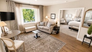 Uma sala de estar moderna com um sofá, mesa de centro e televisão. Há uma janela grande com cortinas, um espelho e vista para um quarto.