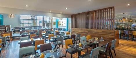 Um restaurante amplo com várias áreas de refeição, um bar e grandes janelas que oferecem iluminação natural.