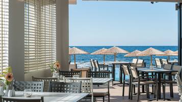 Restaurante à beira-mar com mesas ao ar livre, vista para o oceano e guarda-sóis.