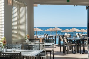 Restaurante à beira-mar com mesas ao ar livre, vista para o oceano e guarda-sóis.