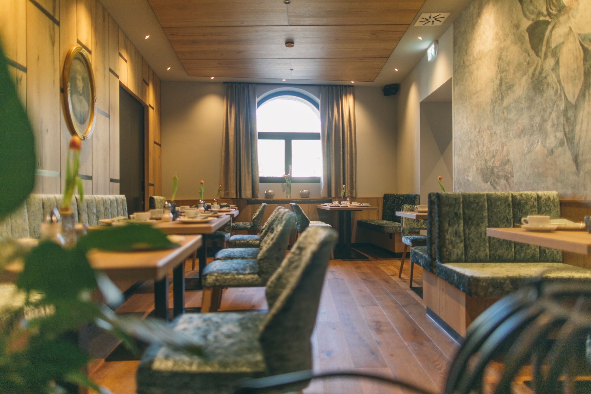 Um restaurante com mesas e cadeiras de madeira, uma janela grande com cortinas e uma parede de mármore com uma escultura.