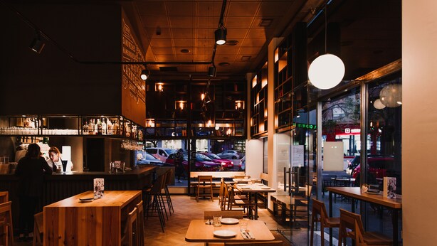 Um restaurante acolhedor com mesas e cadeiras de madeira, um bar e amplas janelas com vista para a rua.
