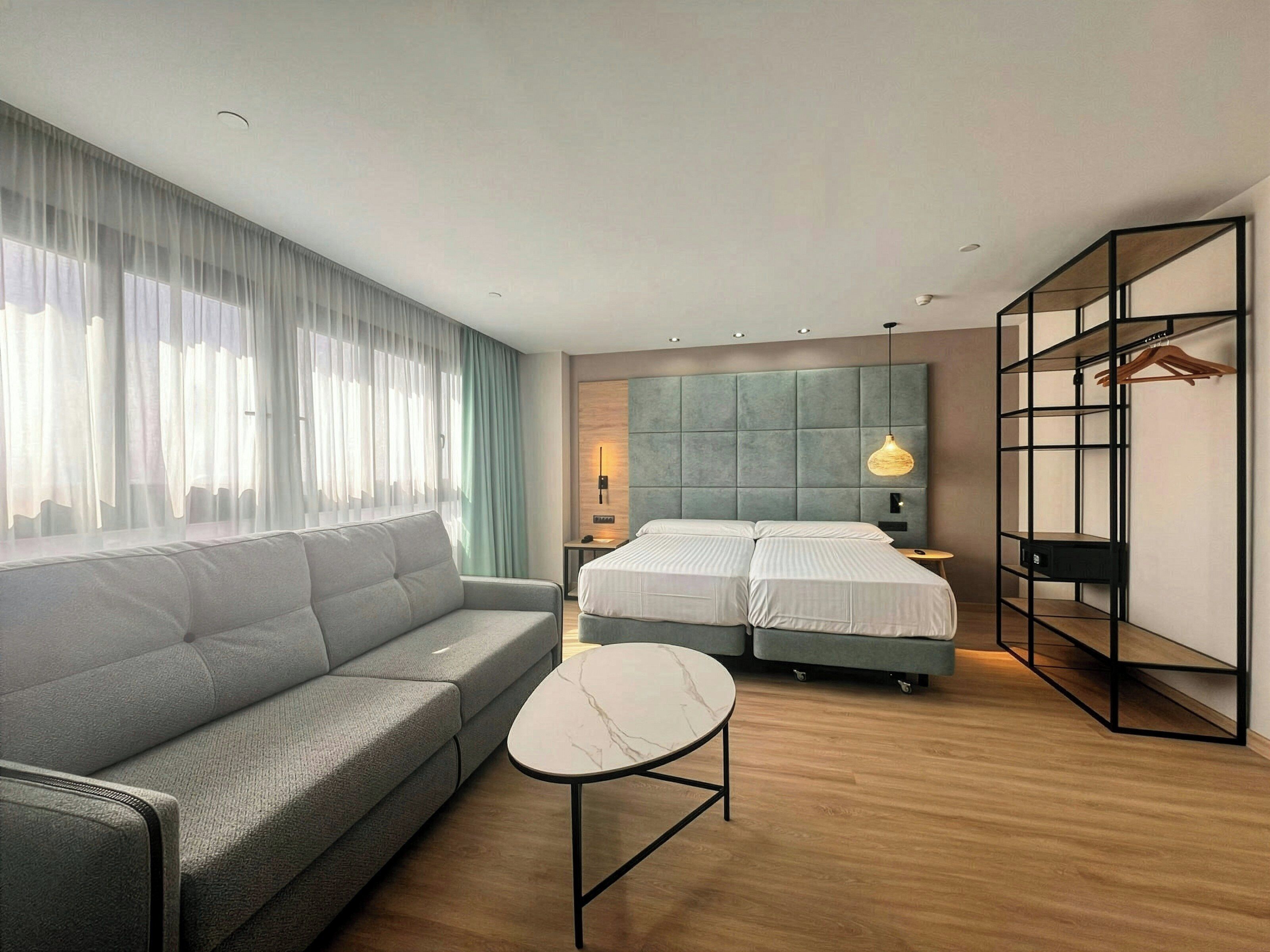 Quarto de hotel moderno com sofá cinza, cama com duas almofadas, mesa redonda pequena e um guarda-roupa com cabideiro.