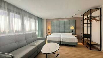 Quarto de hotel moderno com sofá cinza, cama com duas almofadas, mesa redonda pequena e um guarda-roupa com cabideiro.