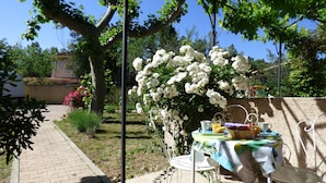 Um pátio externo com uma mesa posta para o café da manhã, cercado por vegetação exuberante e flores brancas.