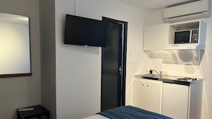 Quarto compacto com cama, televisão na parede, micro-ondas e pequena área de cozinha.
