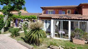 Uma casa tradicional em estilo mediterrâneo, com telhado de telhas, uma estufa de vidro e um jardim com diversas plantas e uma palmeira.