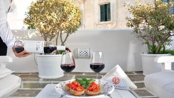 Uma mesa com um prato de bruschetta e duas taças de vinho tinto, uma pessoa servindo vinho, e um edifício ao fundo.