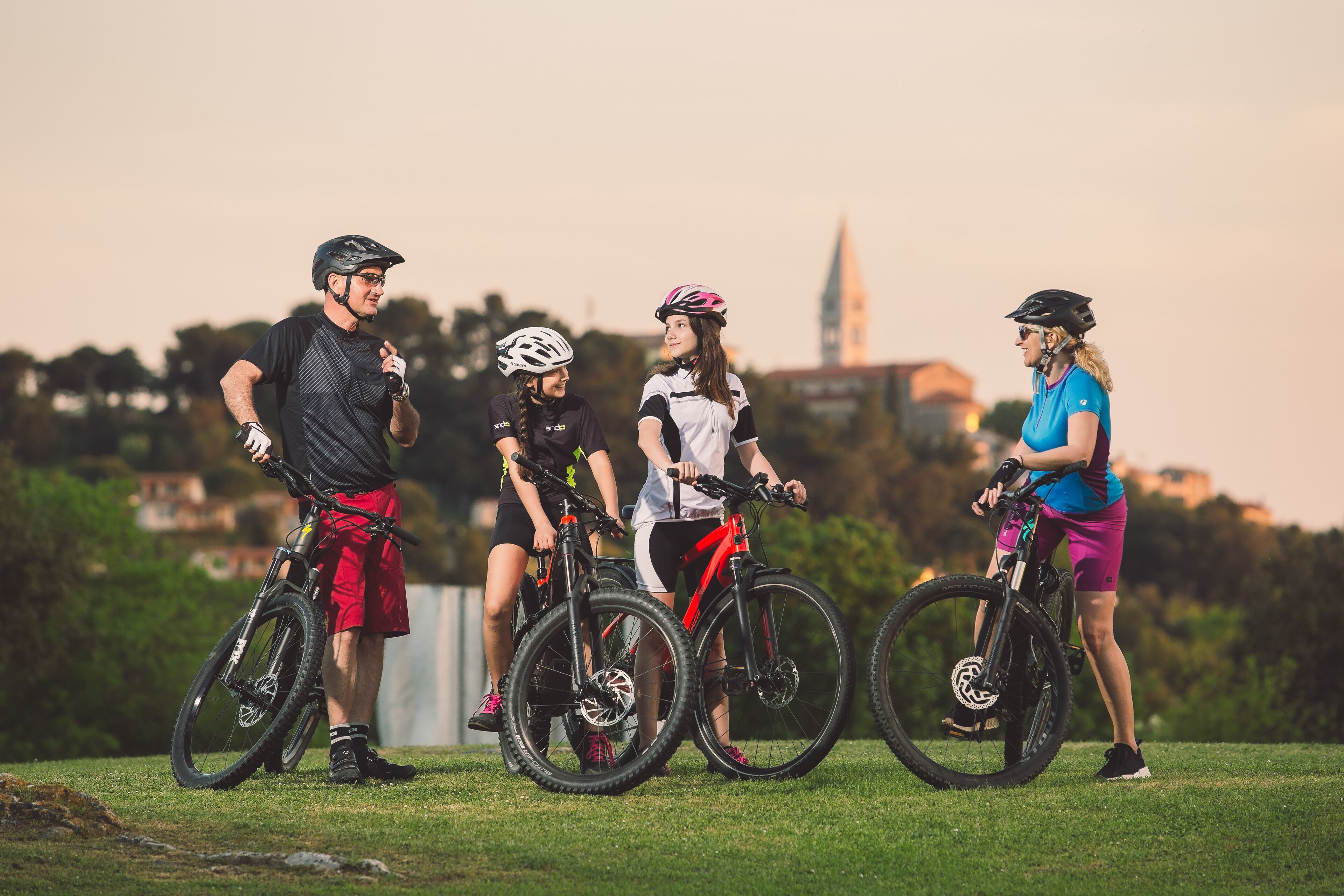Um grupo de pessoas em bicicletas, em ambiente externo, com uma área gramada e um edifício ao longe.