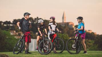 Um grupo de pessoas em bicicletas, em ambiente externo, com uma área gramada e um edifício ao longe.