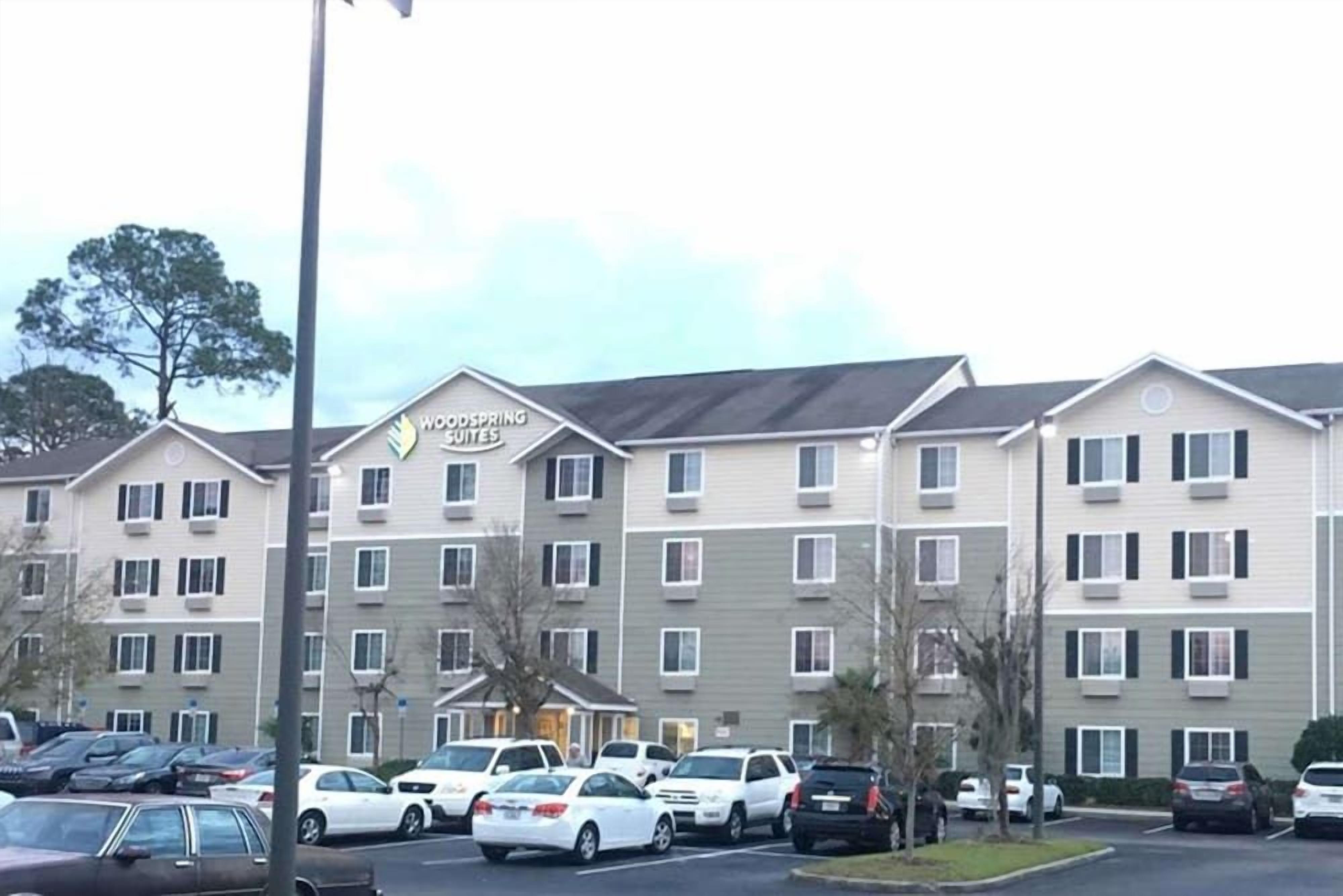 Um hotel com vários andares e uma placa indicando 'Woodspring Suites'. Vários carros estão estacionados no estacionamento, e o edifício apresenta um estilo arquitetônico moderno, com telhado plano e várias janelas.