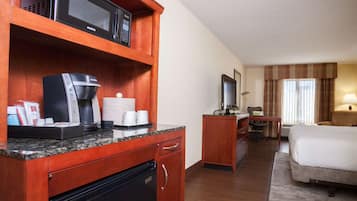 Quarto de hotel com cozinha compacta, incluindo micro-ondas, máquina de café e artigos de higiene. Há uma cama, uma mesa de trabalho com cadeira e uma televisão.