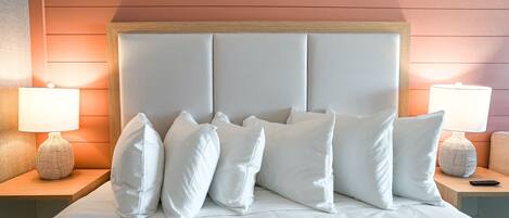 Uma cama bem arrumada com lençóis brancos e várias almofadas, com duas mesas de cabeceira laterais, e um controle remoto em uma delas.