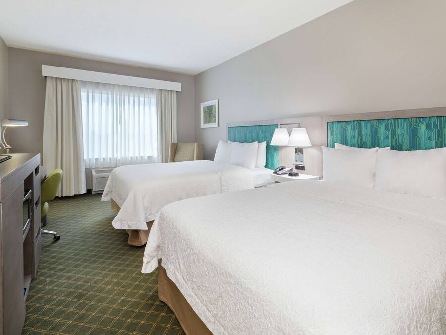 Quarto de hotel com duas camas, uma mesa com televisão, uma cadeira e janela com cortinas.