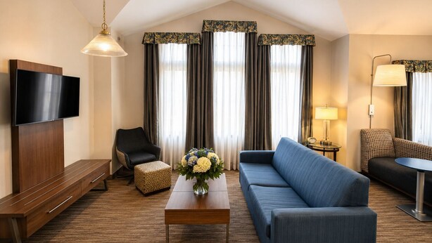 Quarto de hotel com televisão de tela plana, um banco de madeira, um sofá azul, uma mesa pequena com um vaso de flores e uma luminária.