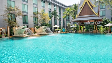 Uma piscina com um pavilhão tradicional tailandês, um edifício moderno com grandes janelas e palmeiras.