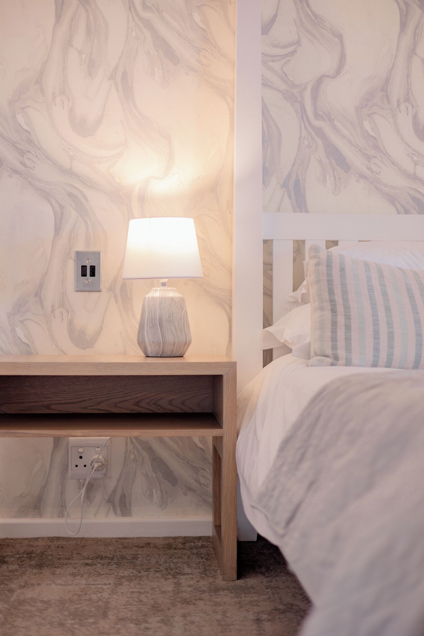 Quarto com parede estampada em mármore, mesa de cabeceira com abajur, tomada e cama com almofadas listradas.