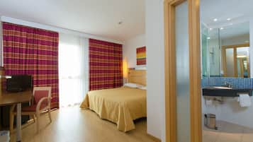 Quarto de hotel com cama, escrivaninha com cadeira, televisão e banheiro com pia e espelho.