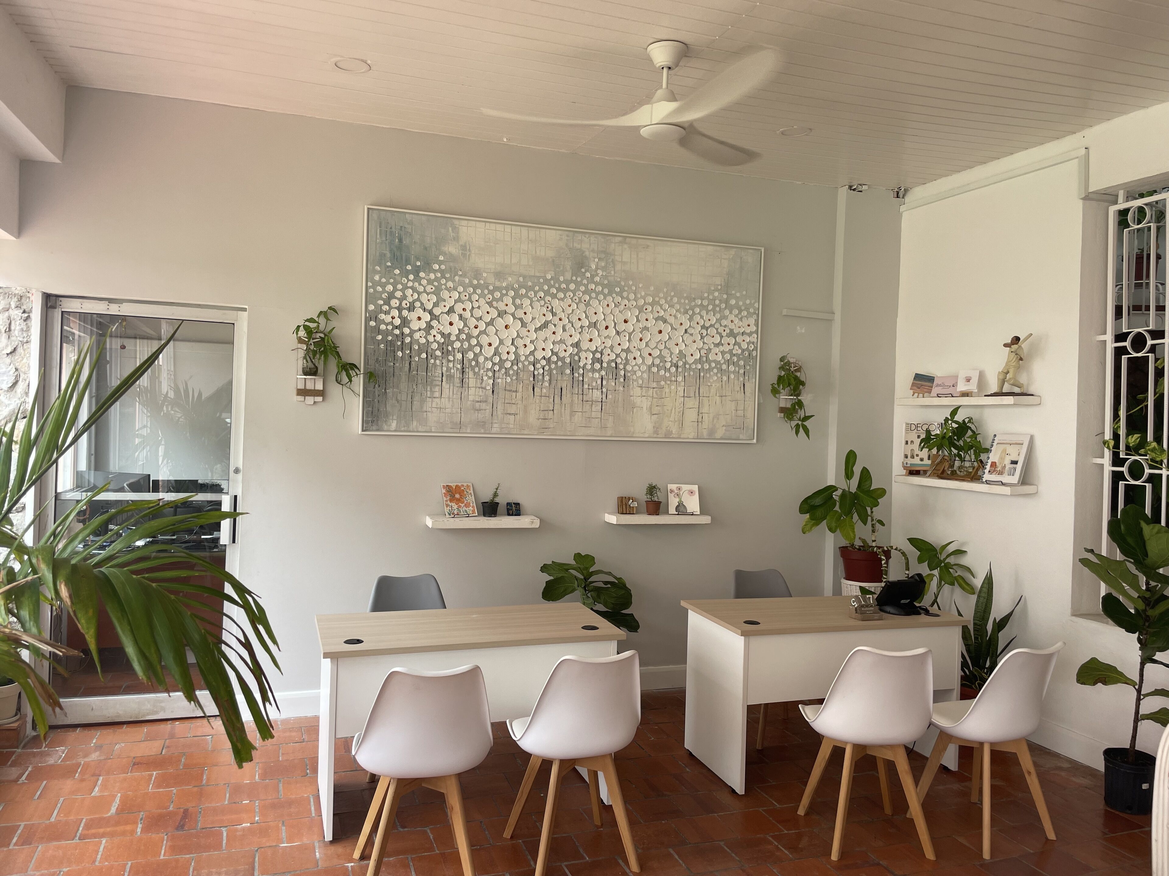 Uma área de jantar moderna com duas mesas brancas e quatro cadeiras brancas, um ventilador de teto e um quadro abstrato grande na parede.