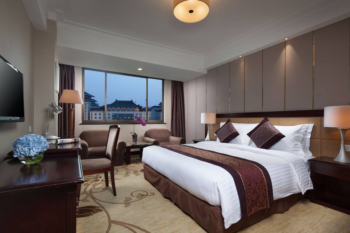 Quarto de hotel com uma cama grande, duas cadeiras, uma escrivaninha e uma televisão.