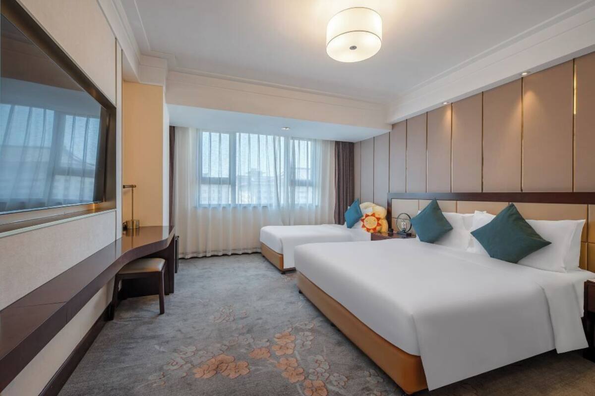 Quarto de hotel com uma cama grande, uma cama menor, uma escrivaninha e uma janela ampla.