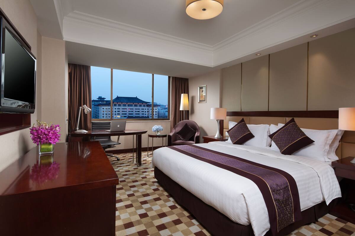 Quarto de hotel com uma cama grande, uma mesa com um laptop, uma TV e vista para a cidade.