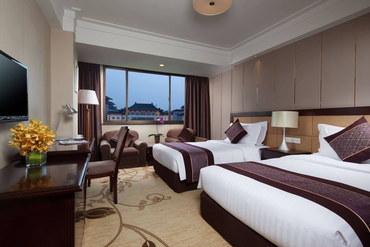 Quarto de hotel com duas camas, uma escrivaninha com televisão, uma cadeira, um abajur e vista para um prédio.