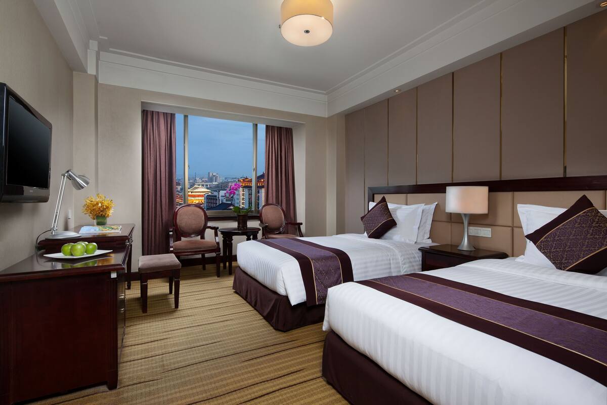 Quarto de hotel com duas camas, uma escrivaninha com luminária, uma cadeira, televisão e vista para a paisagem urbana.