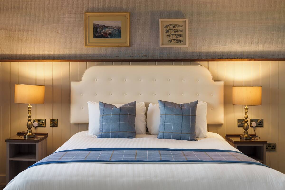 Quarto de hotel com uma cama grande, duas mesas de cabeceira com luminárias, cabeceira com botões e quadros na parede.