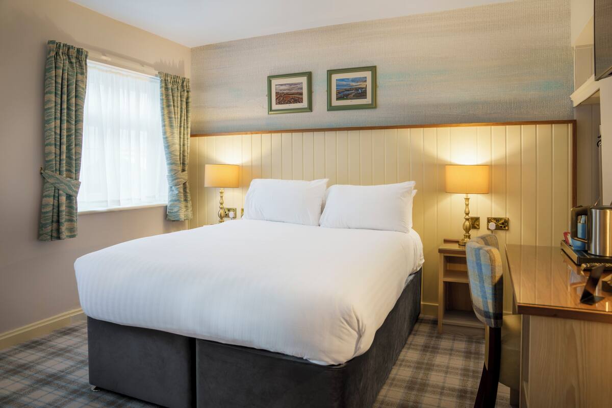 Quarto de hotel com uma cama grande, duas luminárias de cabeceira, uma escrivaninha com cadeira e uma janela com cortinas.
