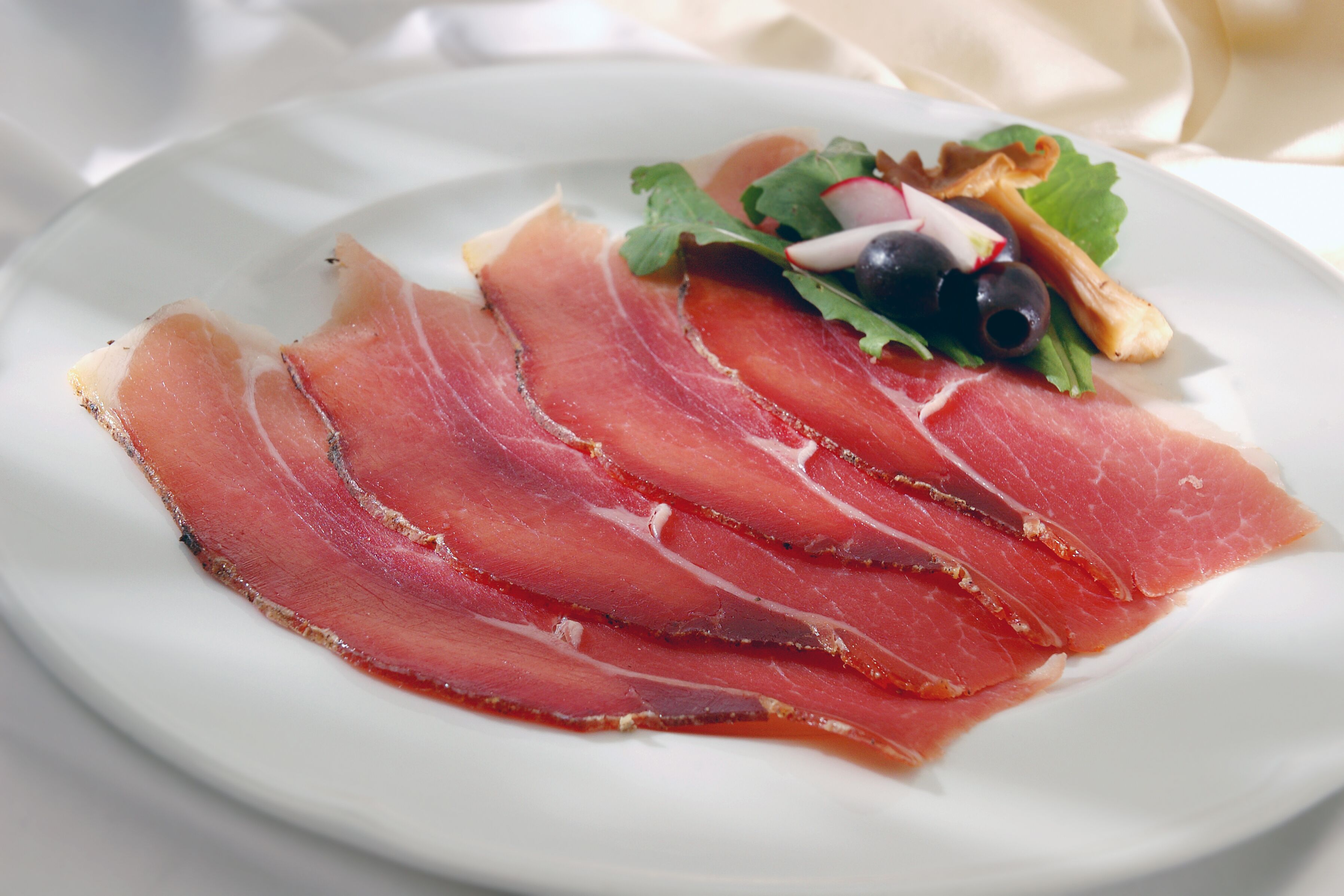 Um prato com fatias finas de carne curada, possivelmente presunto, acompanhado de uma salada pequena com folhas, rabanetes e azeitonas.
