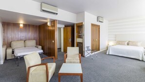 Quarto de hotel com uma cama, um sofá, uma cadeira, uma mesinha e uma secretária. Há um guarda-roupa de madeira e uma porta que dá para outro quarto.