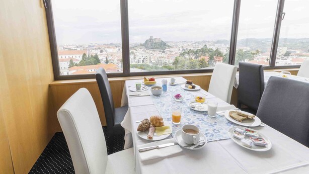Mesa de pequeno-almoço com pratos de comida, uma chávena de café e vista para a paisagem urbana através de grandes janelas.