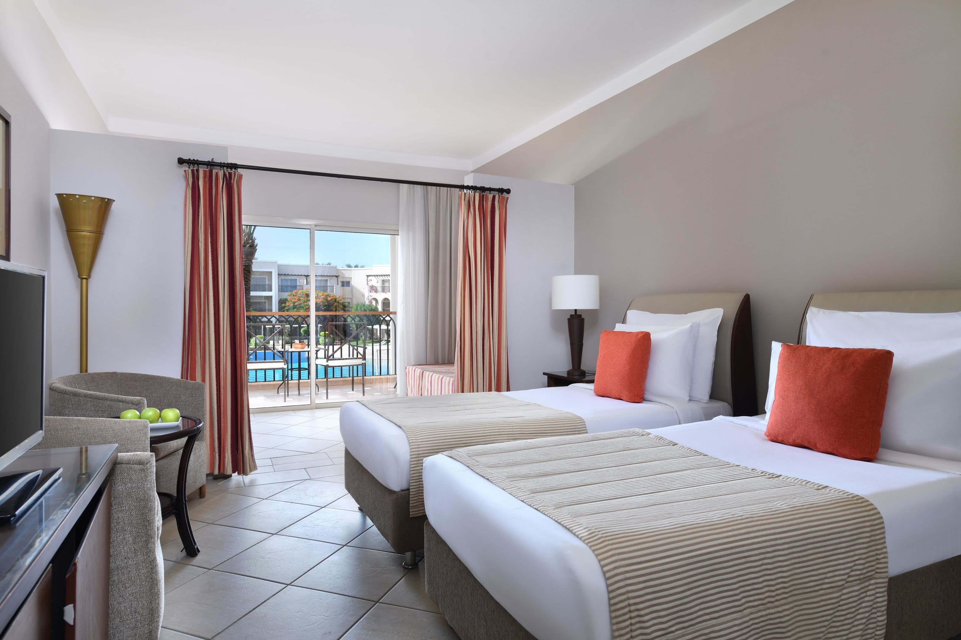 Quarto de hotel com duas camas, televisão, escrivaninha com cadeira, luminária e vista para uma piscina.