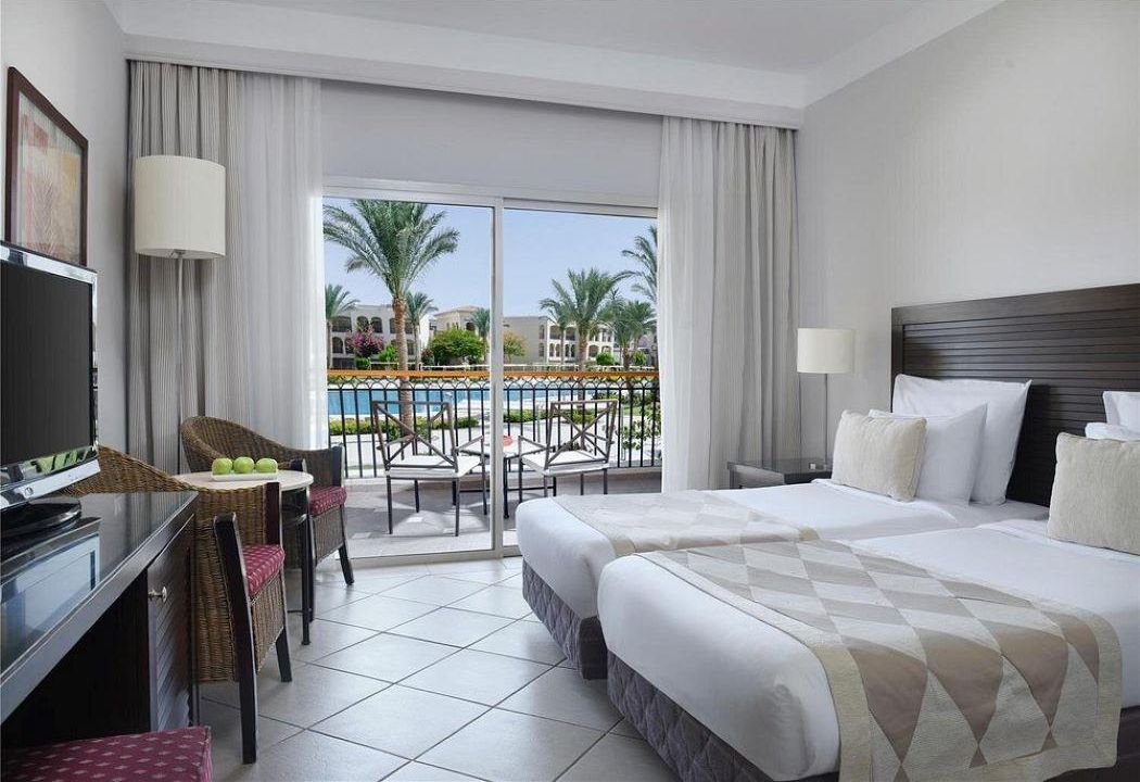 Quarto de hotel com cama, escrivaninha, cadeira, televisão e varanda com vista para palmeiras e piscina.