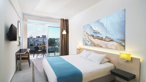 Quarto de hotel com cama, escrivaninha e cadeira; uma janela grande com vista para edifícios; e um quadro na parede.