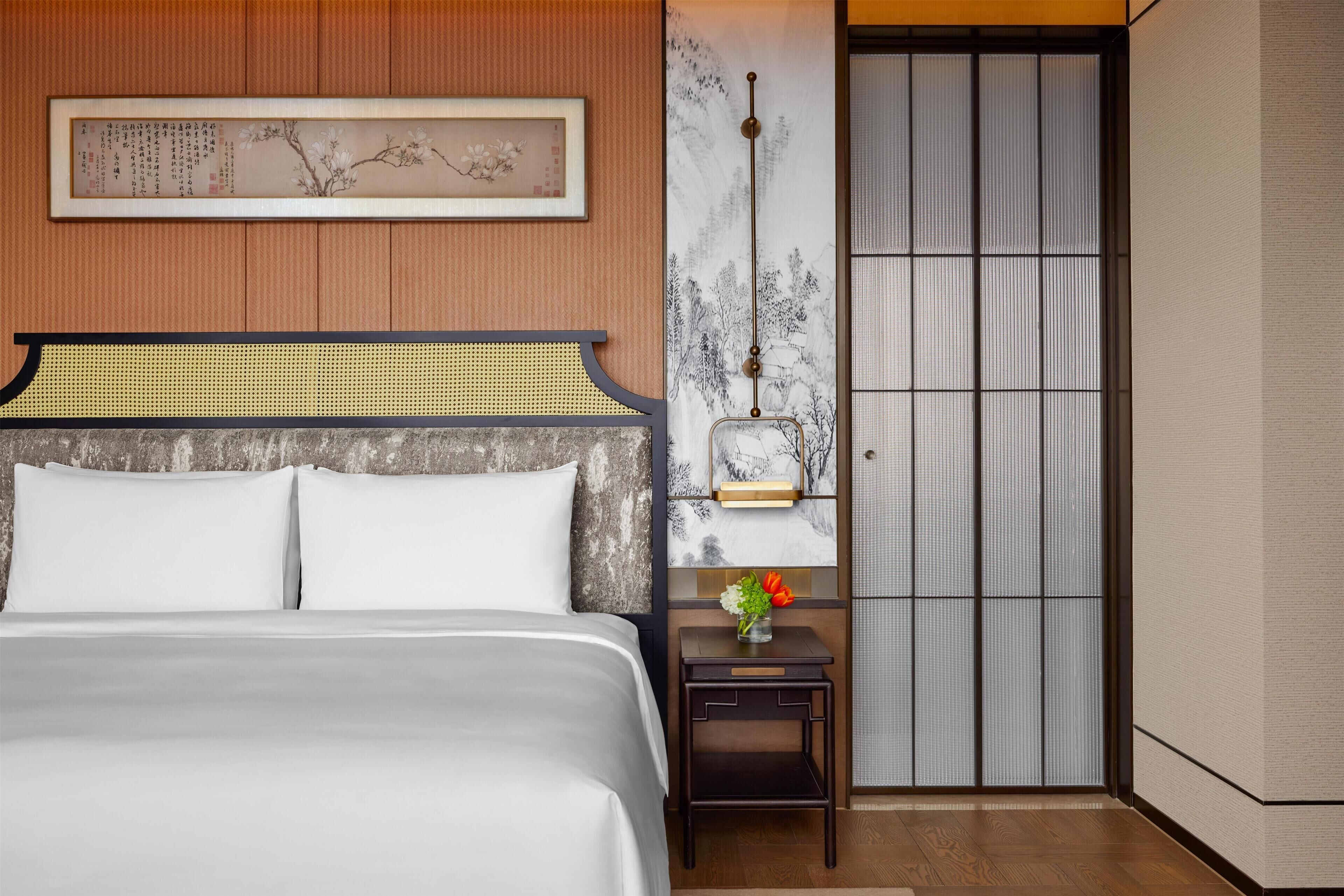 Quarto de hotel com cama, mesa de cabeceira com um vaso de flores, uma obra de arte emoldurada na parede e uma porta de correr com vista.