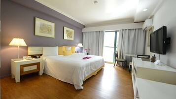 Quarto de hotel com uma cama grande, mesas de cabeceira, escrivaninha, cadeira, televisão e janela com cortinas.
