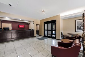 Uma área de recepção de hotel com balcão, uma tela digital exibindo 'Wi-Fi Grátis em Todos os Quartos' e um espaço de estar com cadeiras e mesa.
