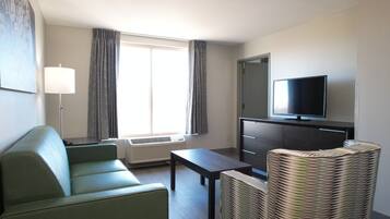 Quarto de hotel pequeno com um sofá verde, uma mesinha, uma televisão em um armário e uma janela com cortinas.