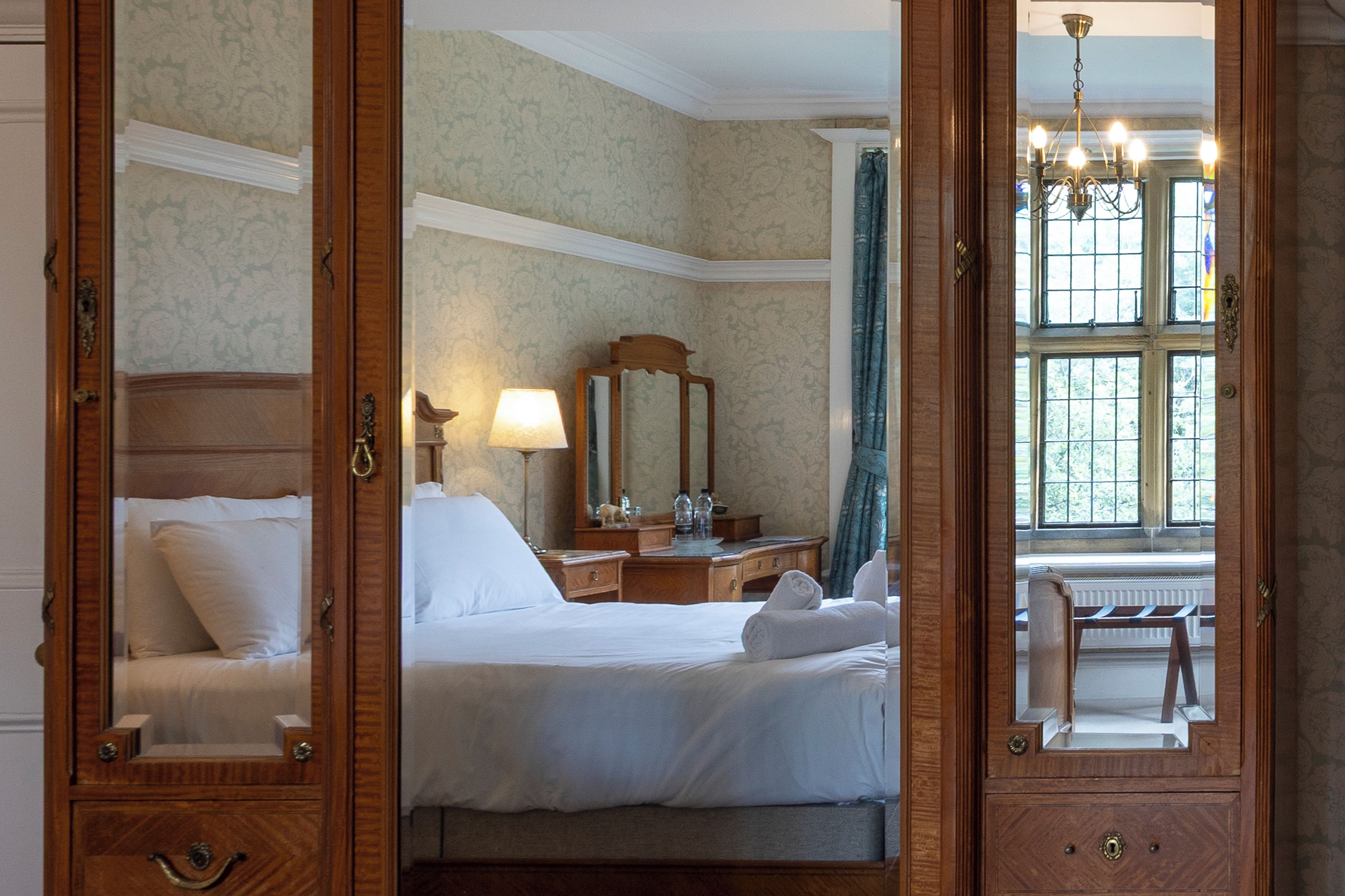 Quarto de hotel com uma cama grande, uma mesa de cabeceira com abajur, um espelho e vista para o exterior pela janela.