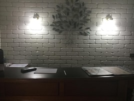Quarto com parede de tijolos, duas luminárias na parede, uma escrivaninha com papéis, uma caneta e uma planta.